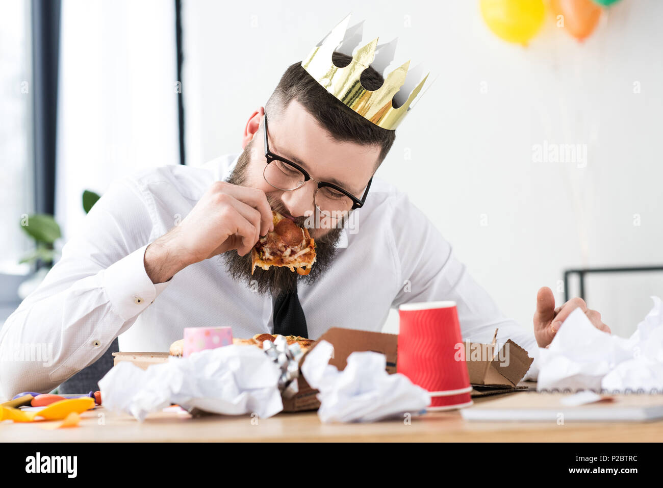 portrait of businessman with paper crown on head eating pizza at ...