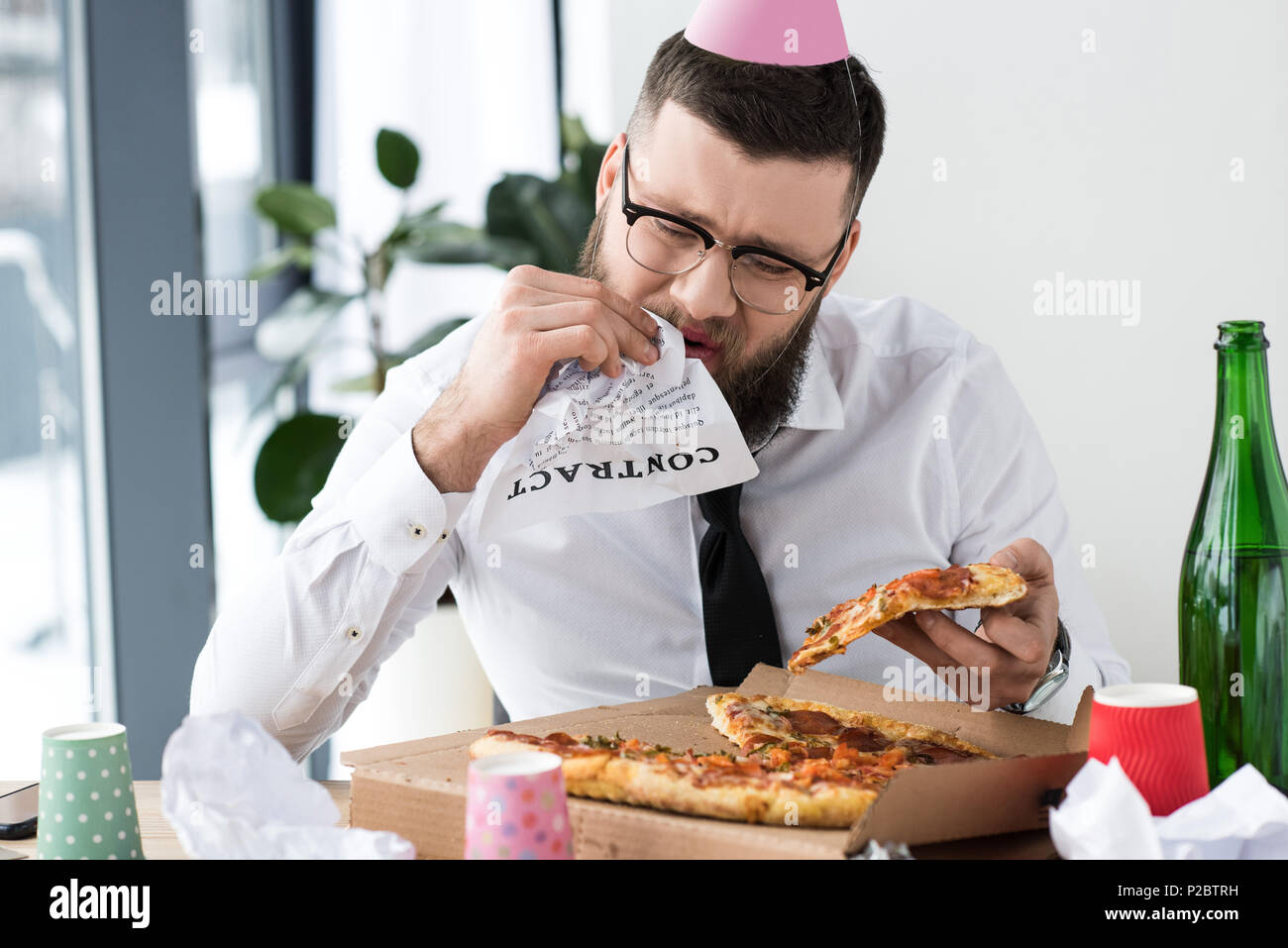 portrait of businessman with party cone on head eating pizza at ...