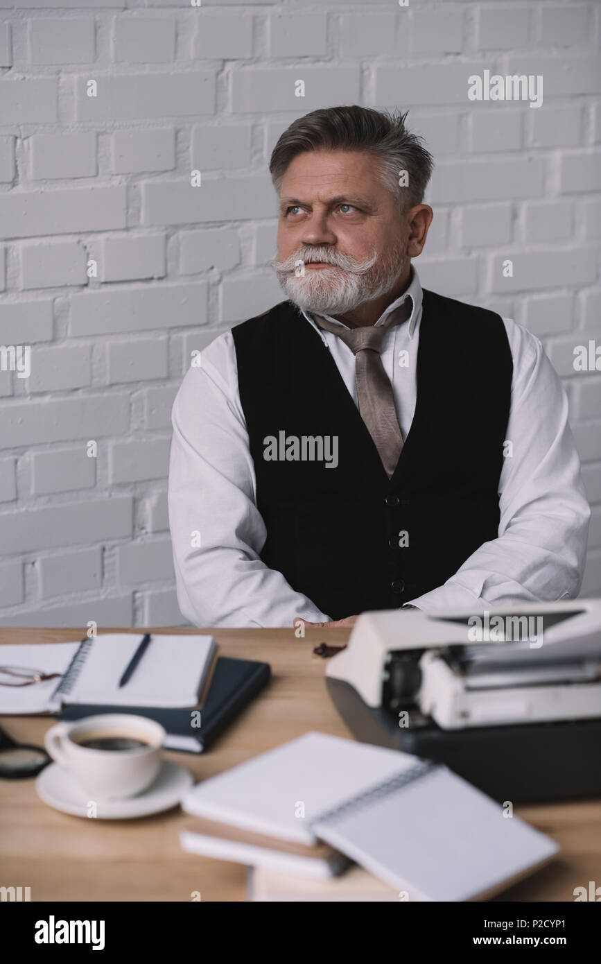 thoughtful senior writer sitting at workplace against white brick wall ...