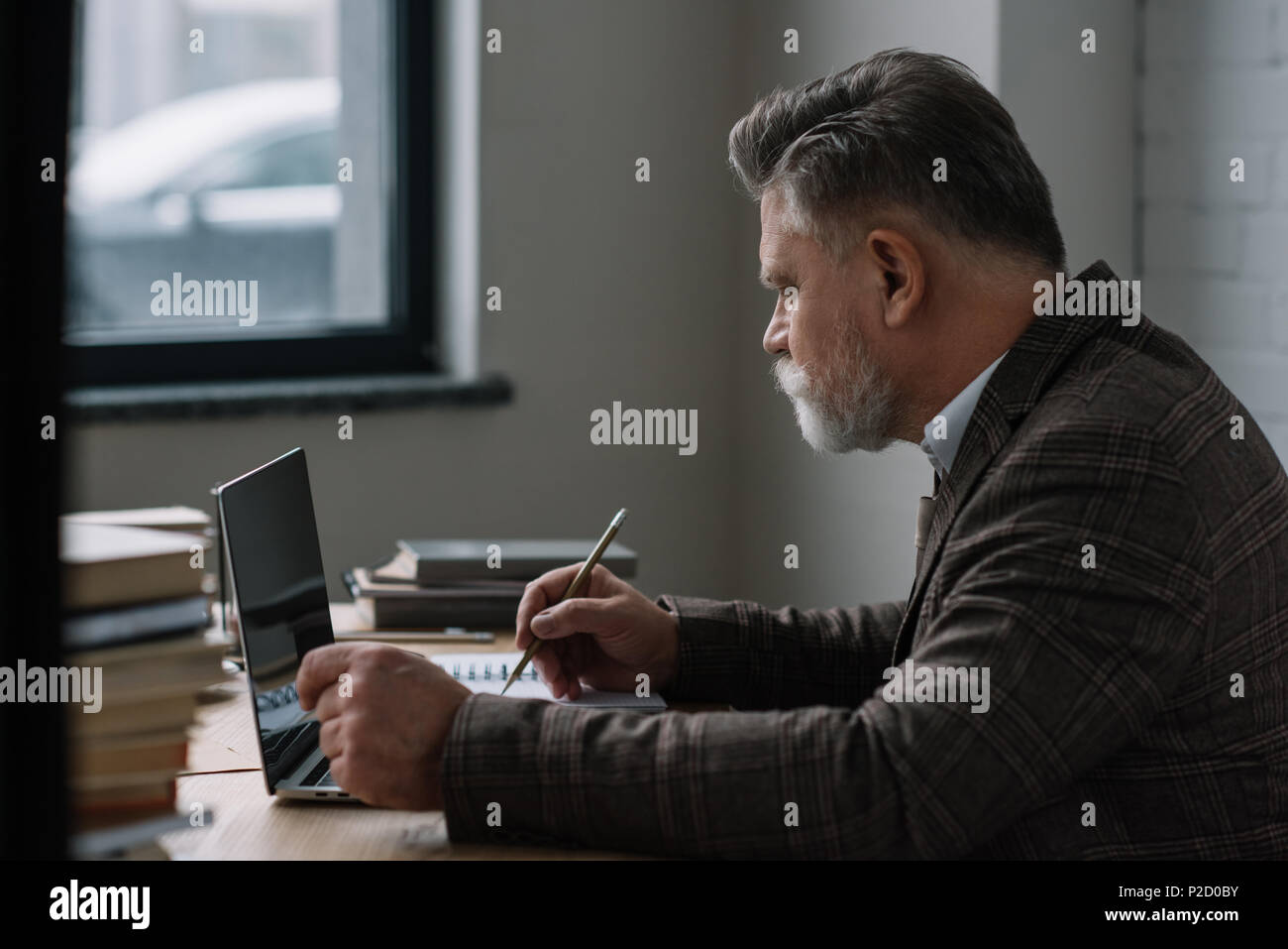 thoughtful senior writer working with laptop and making notes in ...