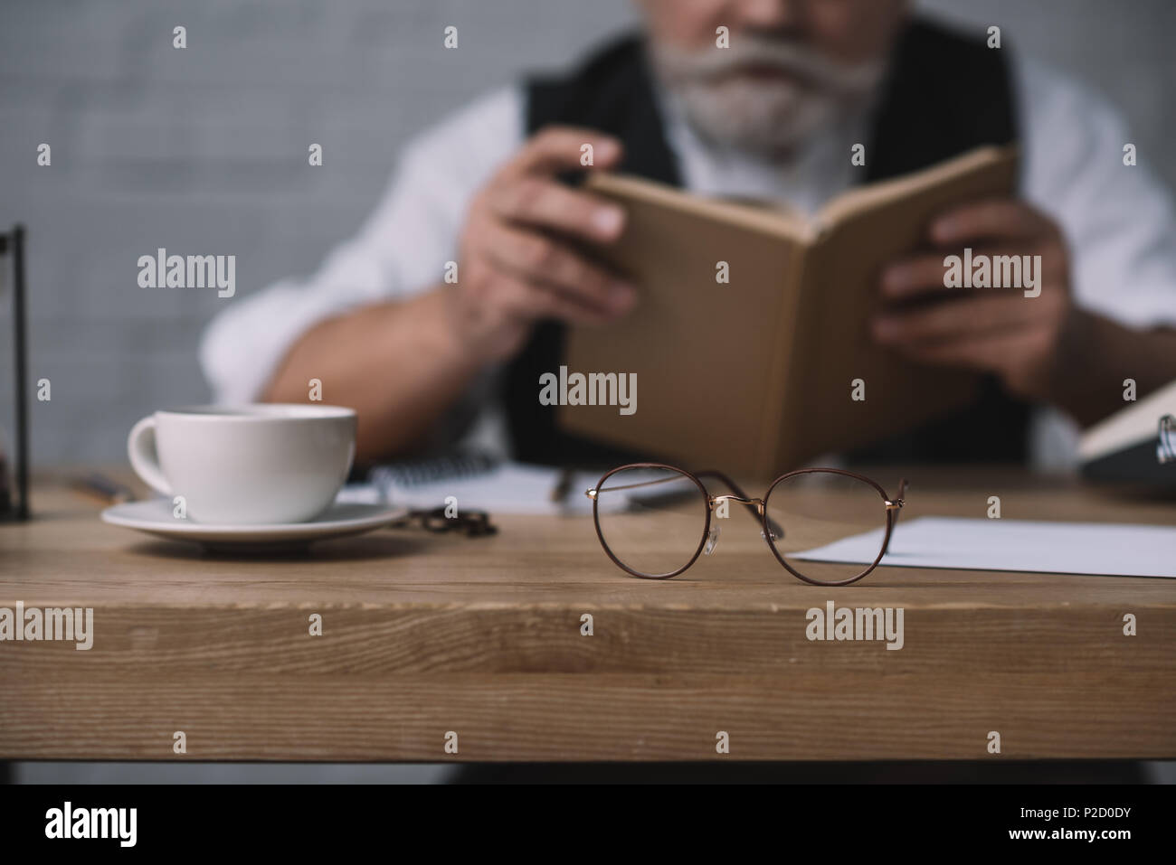 senior man reading book at work desk with cup of coffee and eyeglasses ...