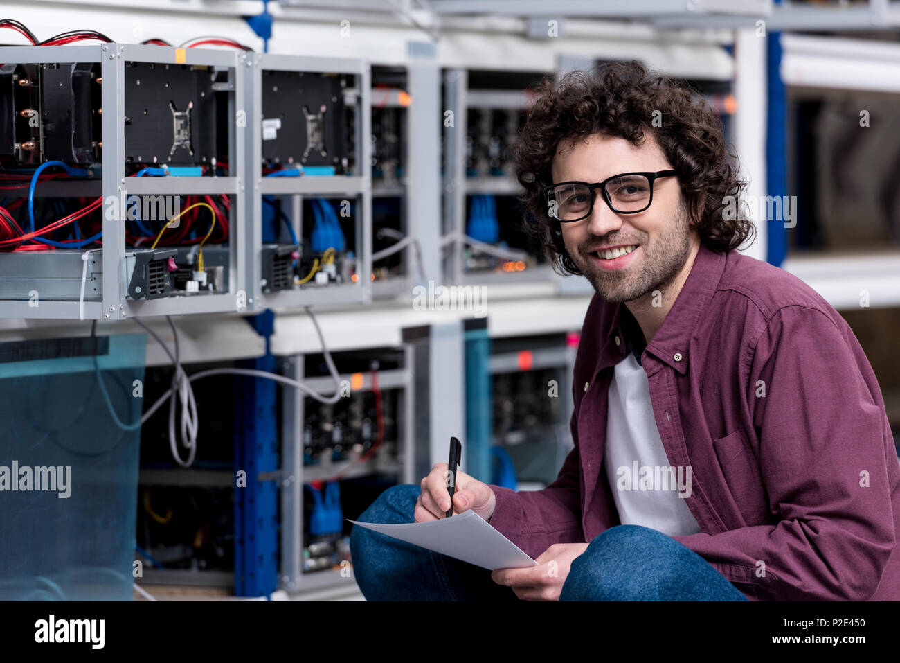 young smiling computer engineer sitting on floor and writing at ...