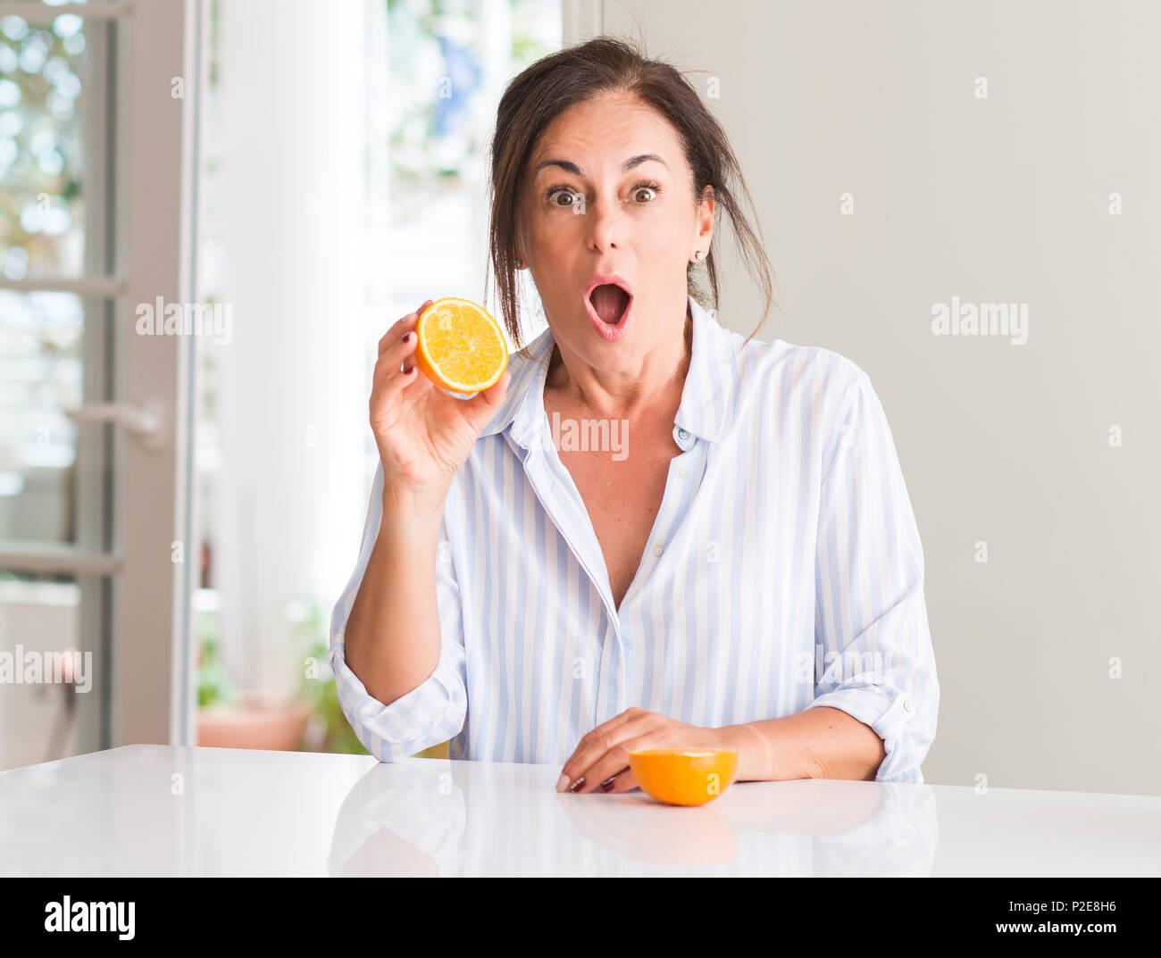 Middle aged woman holding orange fruit scared in shock with a surprise ...