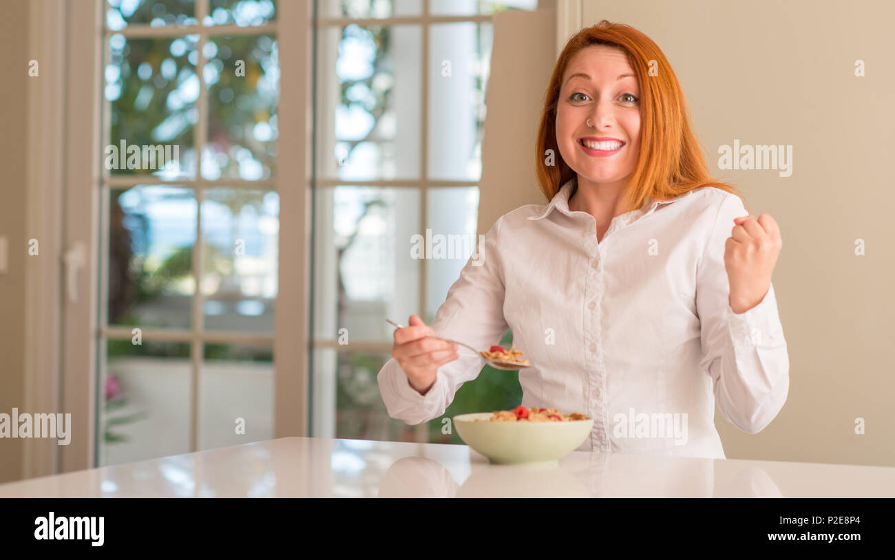 Redhead woman eating cereals with raspberries at home screaming proud ...