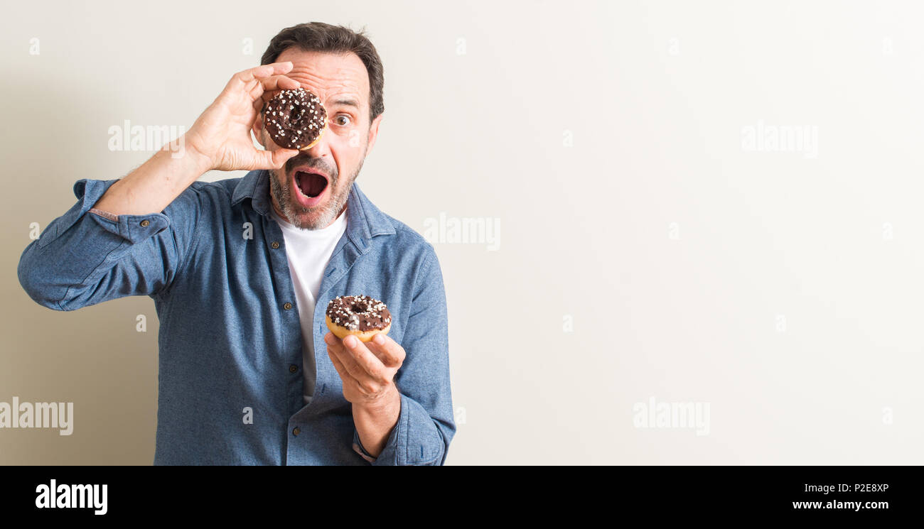 Senior man eating chocolate donut scared in shock with a surprise face ...