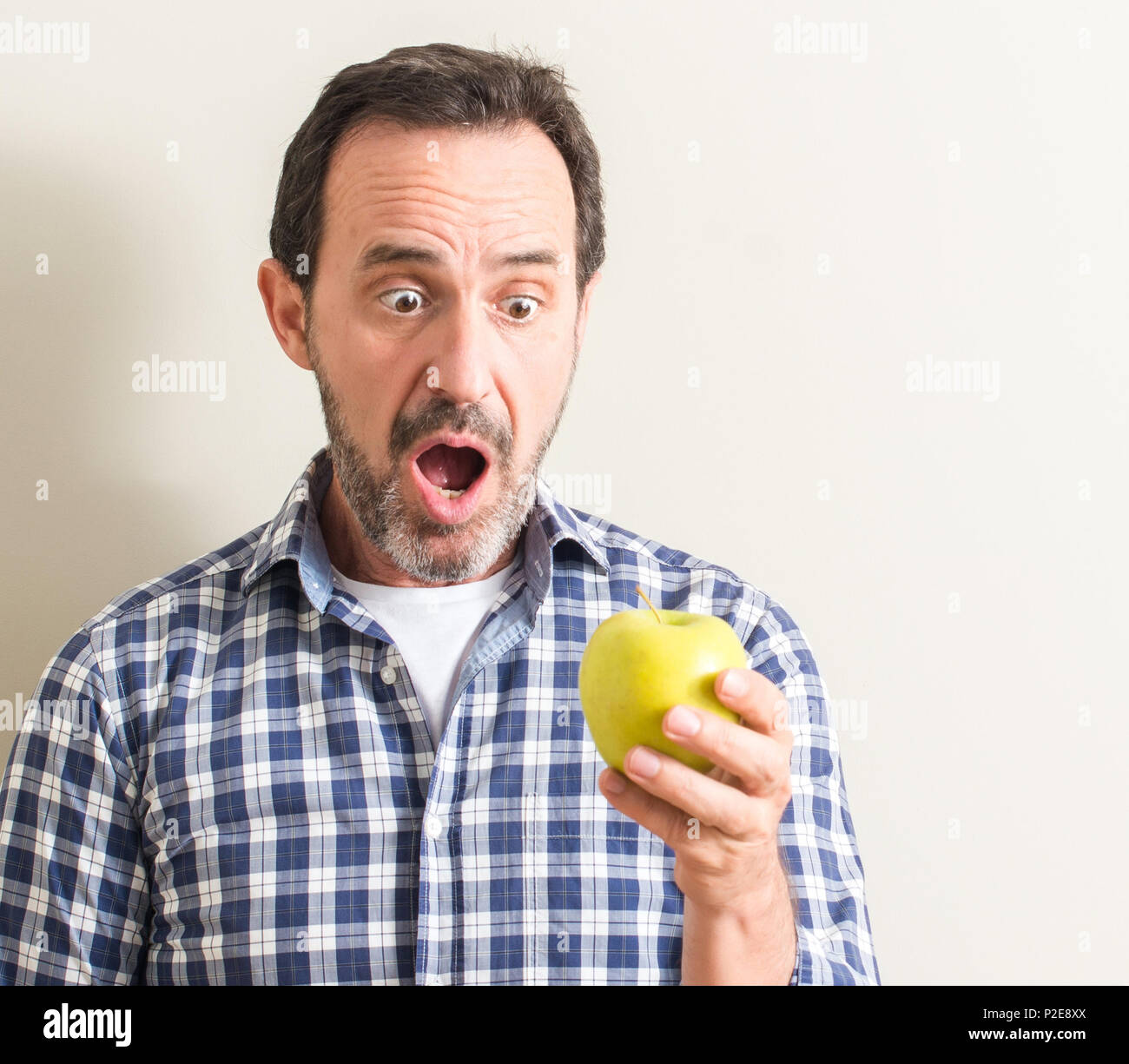 Senior man holding a green apple scared in shock with a surprise face ...