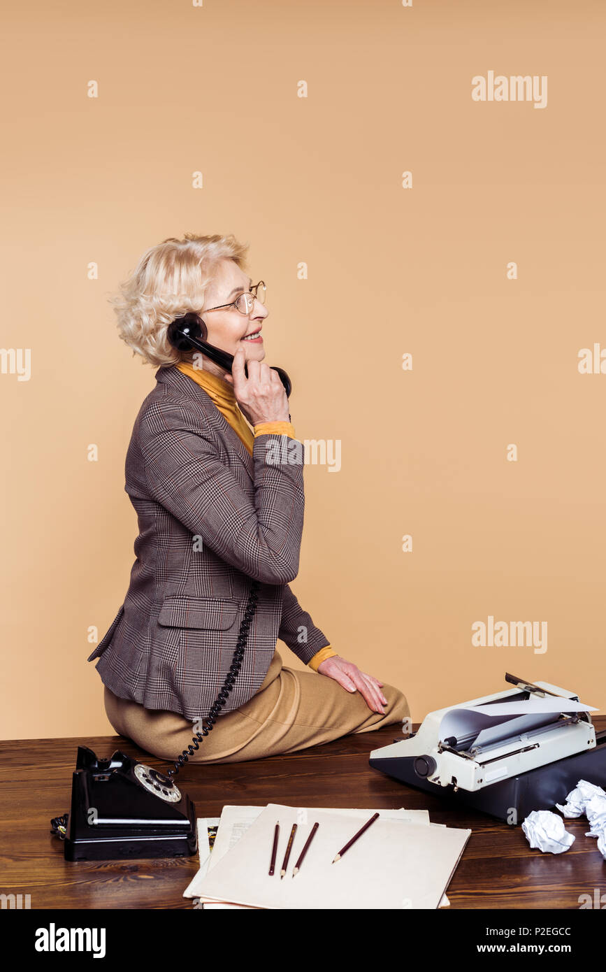 side view of smiling senior woman talking on rotary phone at table with ...