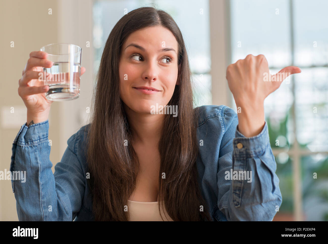 Young woman drinking glass of water at home pointing with hand and ...