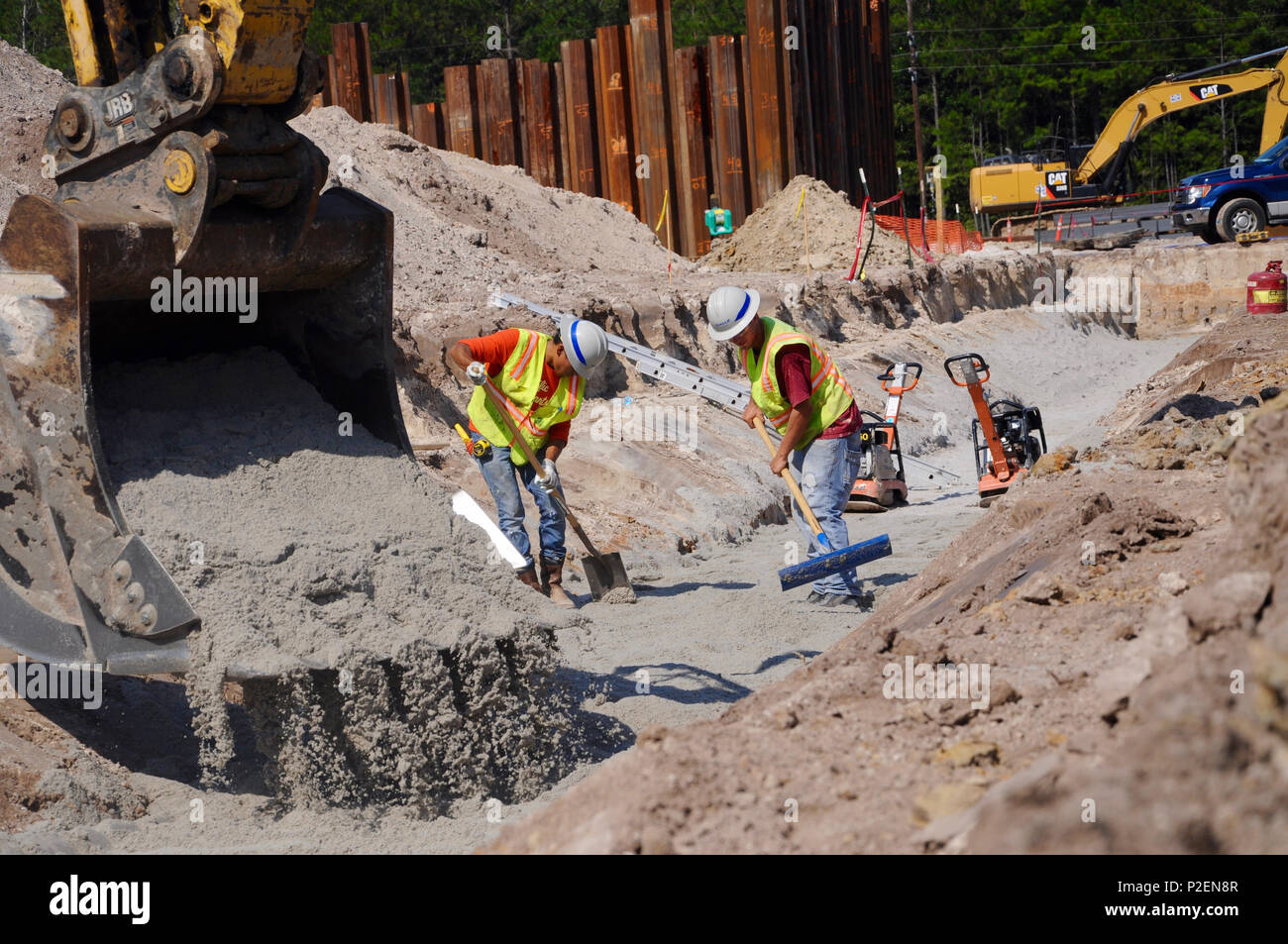 A backhoe loader fills a toe drain with coarse sand as two workers from ...