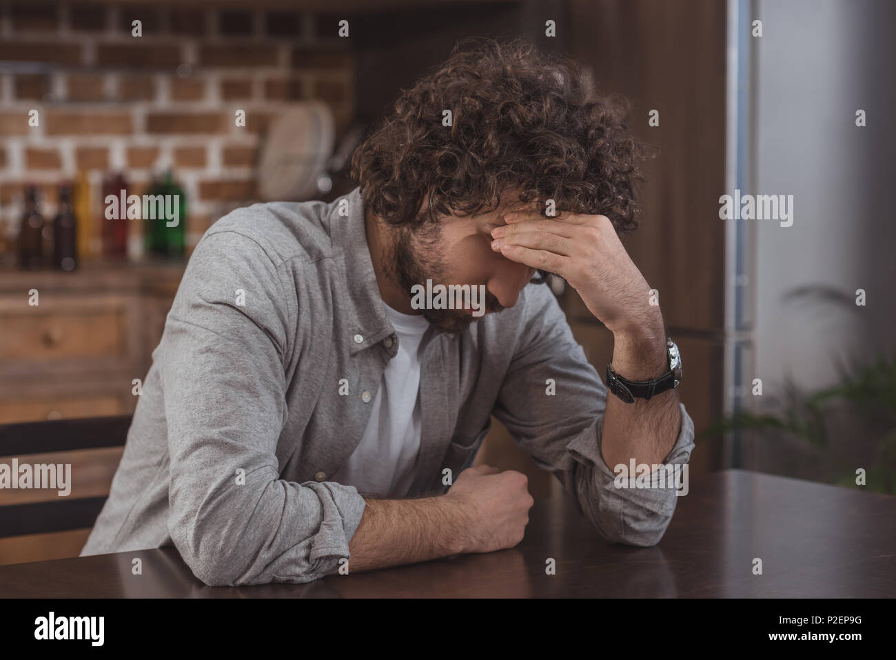 tired man sitting at table in kitchen and touching head Stock Photo - Alamy