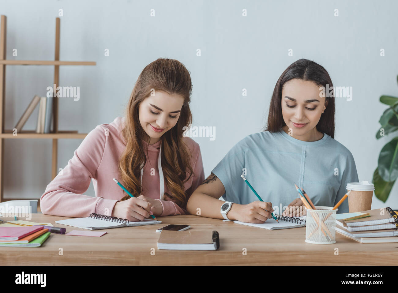 multiethnic girls studying together and writing in copybooks at table ...