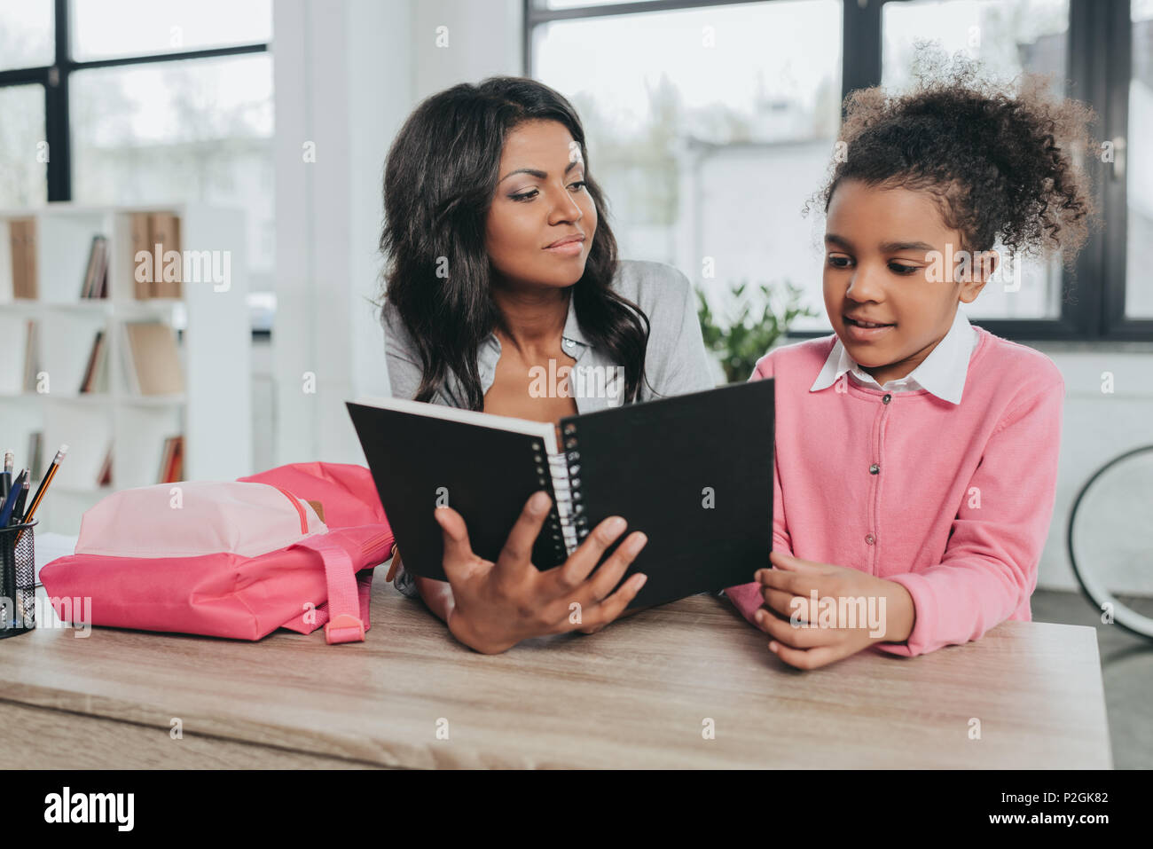 Smiling mother and daughter sitting at wooden table and reading ...