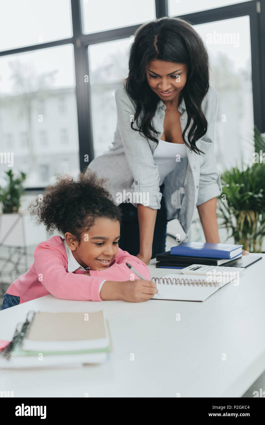 smiling mother looking at little daughter writing in notebook at ...