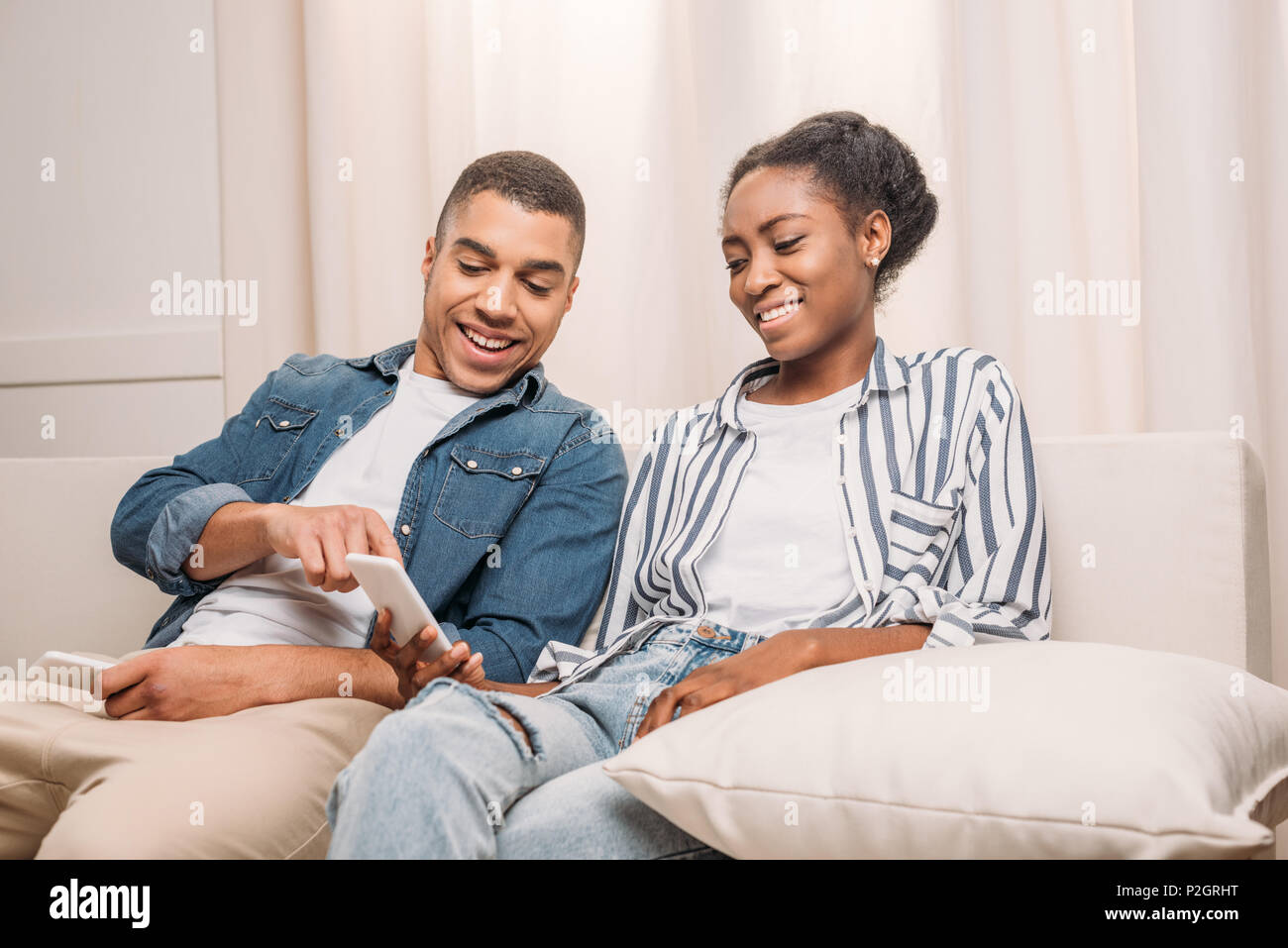 young african american couple sitting on couch with smartphones and ...