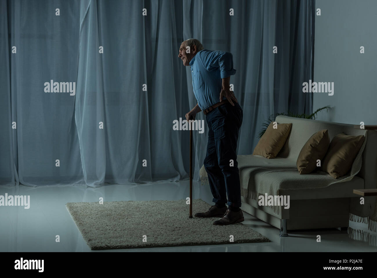 Senior man suffering from back pain leans on a cane in empty room ...