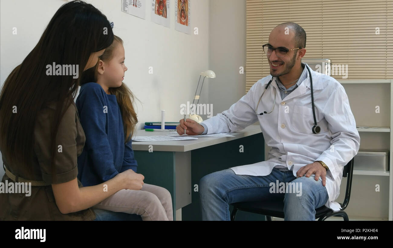 Smiling male doctor talking with shy little girl sitting on her mother ...