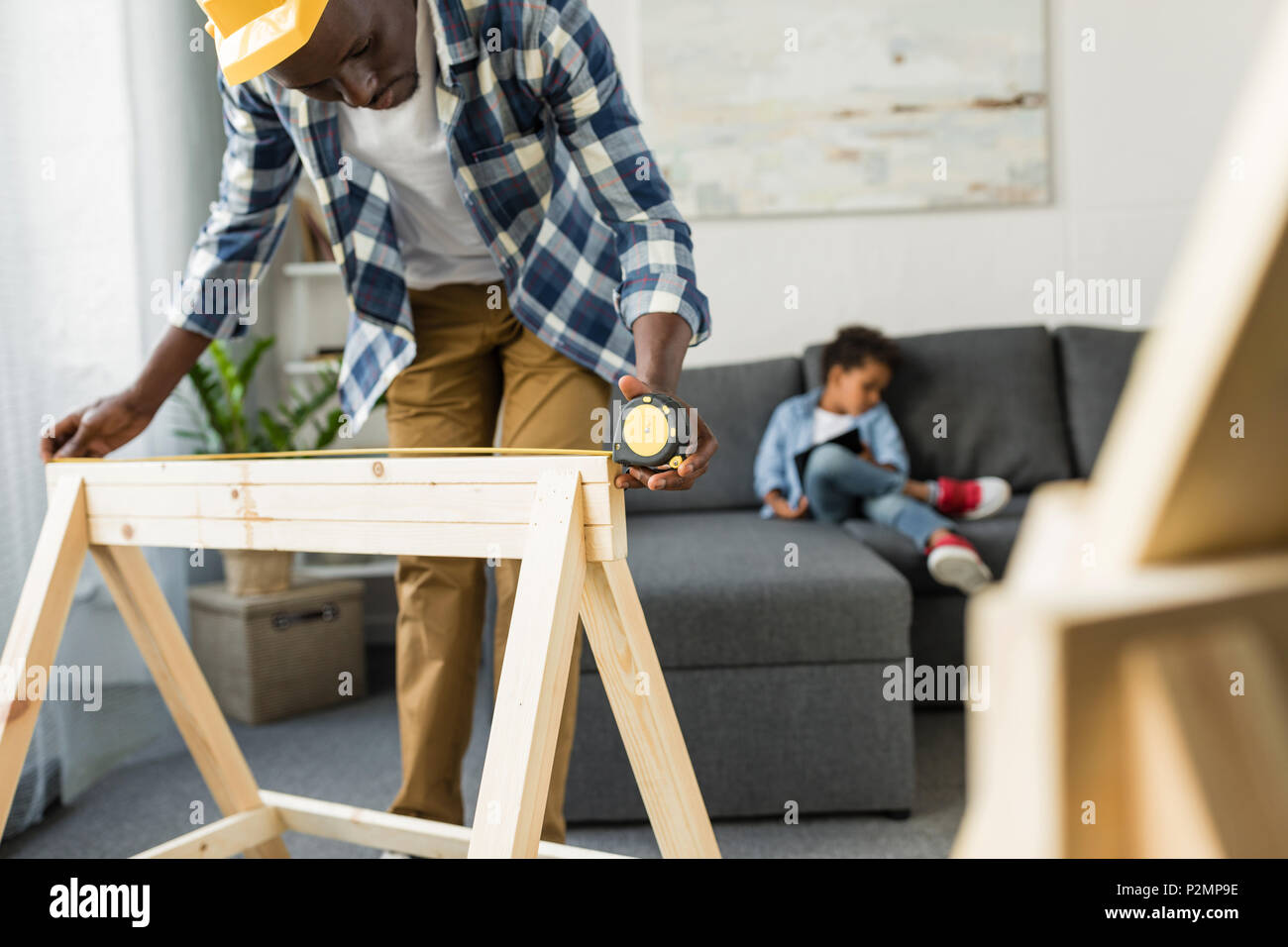 man measuring wooden plank with tape while his son sitting on sofa ...