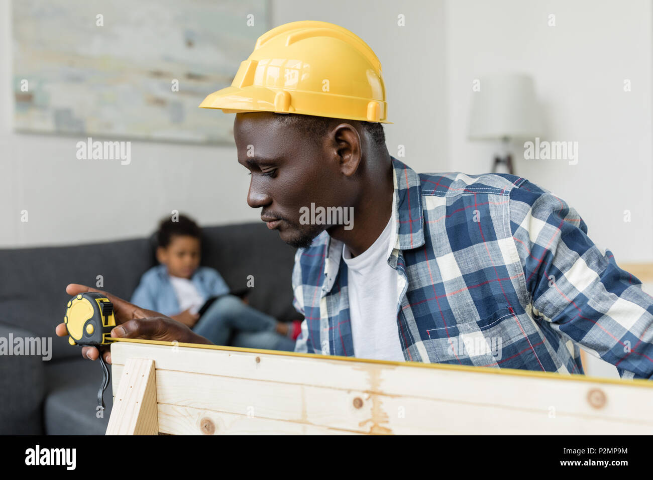 man measuring wooden plank with tape while his son sitting on sofa ...