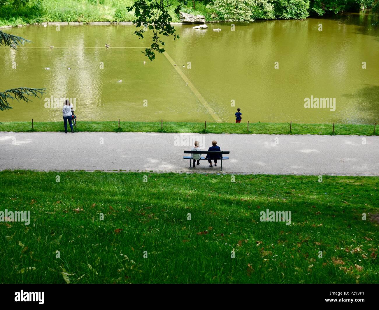 Two women sit on a bench overlooking the lake at Parc Buttes Chaumont ...