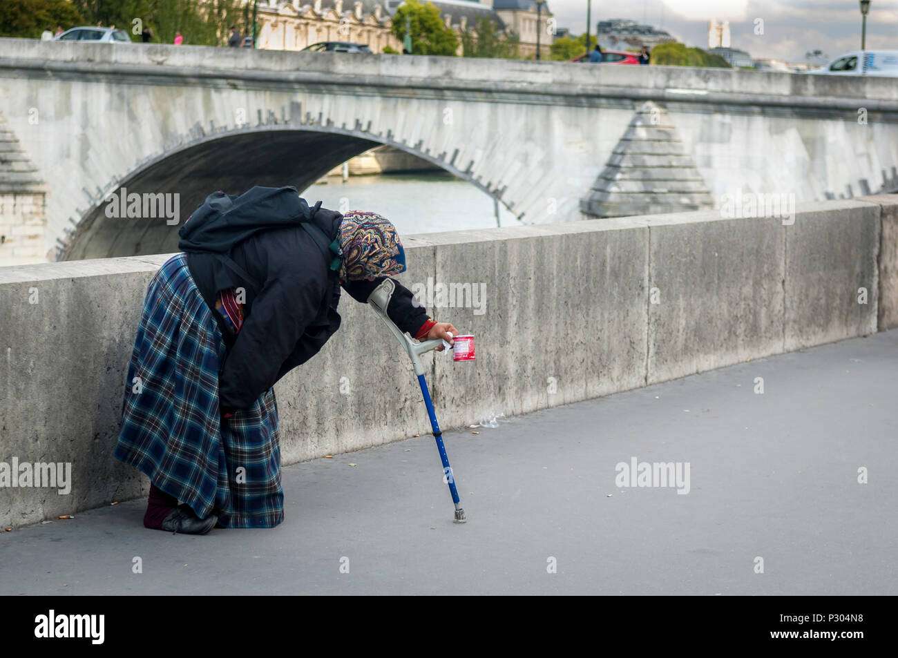 An elderly homeless woman, leaning on a walking stick for support, near ...