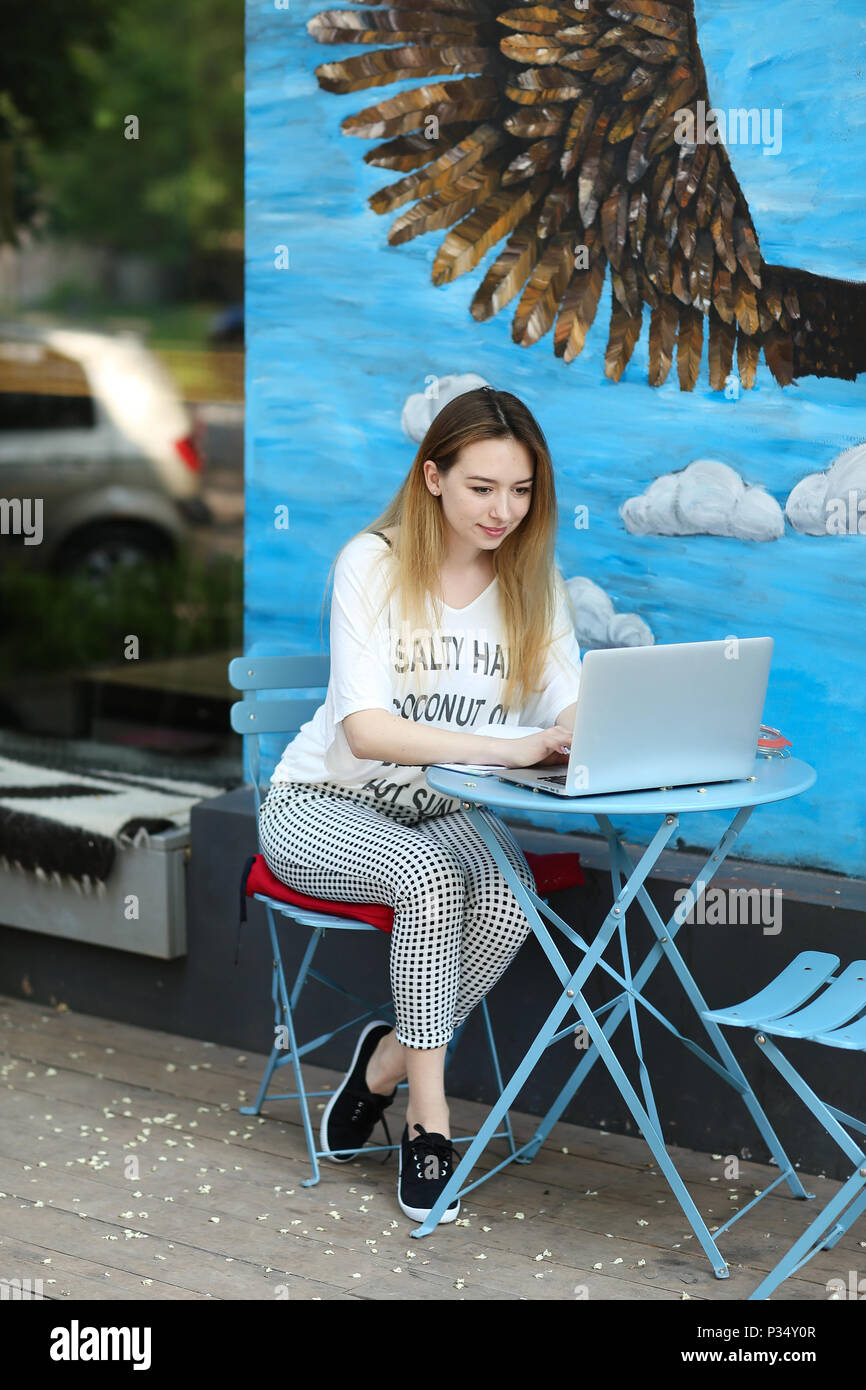Young blonde woman working with laptop at street cafe near painted wall ...
