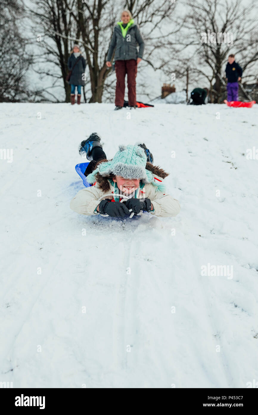 Little girl is lying on her belly on a sled, zooming down a big hill in ...