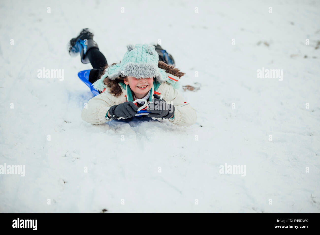 Little girl is lying on her belly on a sled, going down a big snowy ...