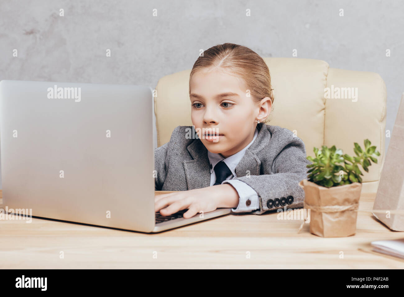 portrait of kid in suit typing on laptop at workplace isolated on grey ...