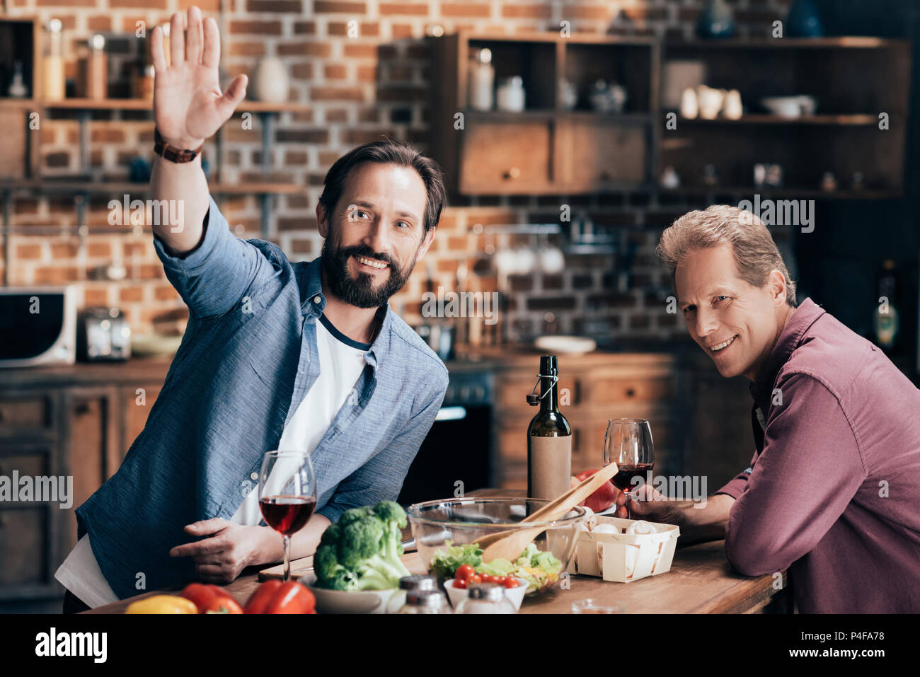 cheerful middle aged men drinking wine and waving hand while cooking ...