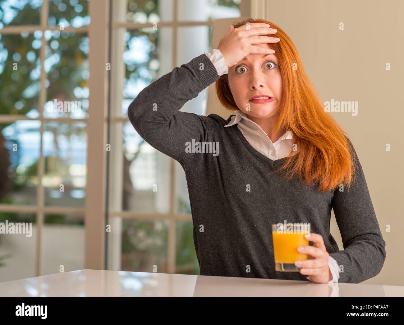 Redhead woman holding orange juice glass at home, stressed with hand on ...