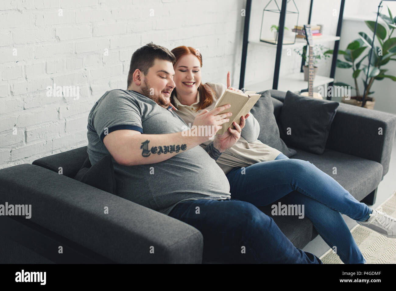 Smiling overweight boyfriend and girlfriend reading book on sofa at ...