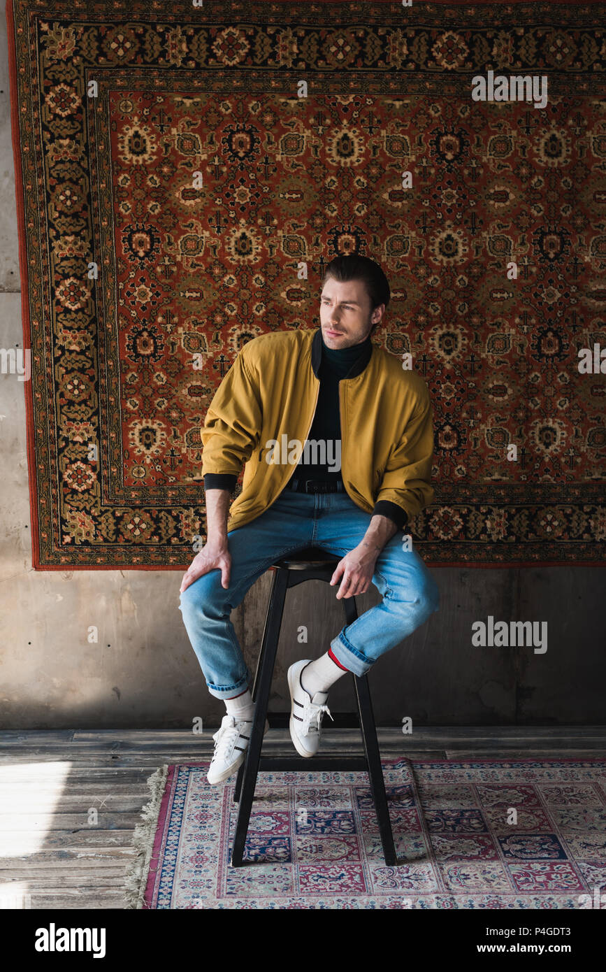 stylish young man sitting on chair in front of rug hanging on wall ...