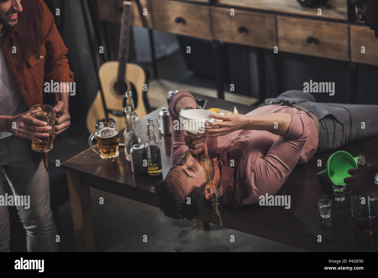 young man lying on table and drinking beer from funnel while friends ...