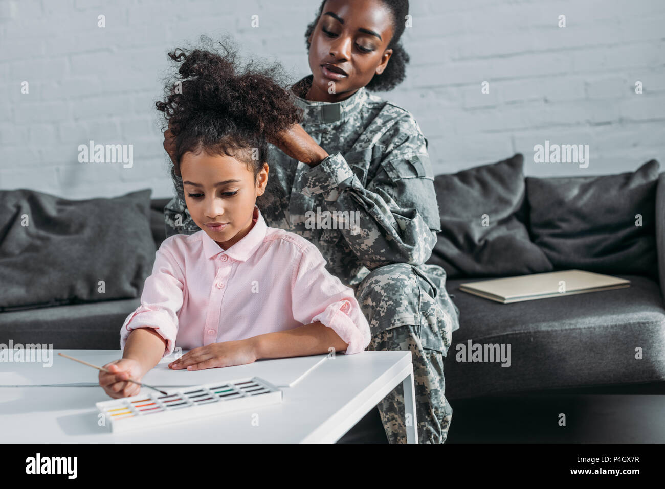 Mother soldier fixing hairstyle of daughter busy with drawing by table ...