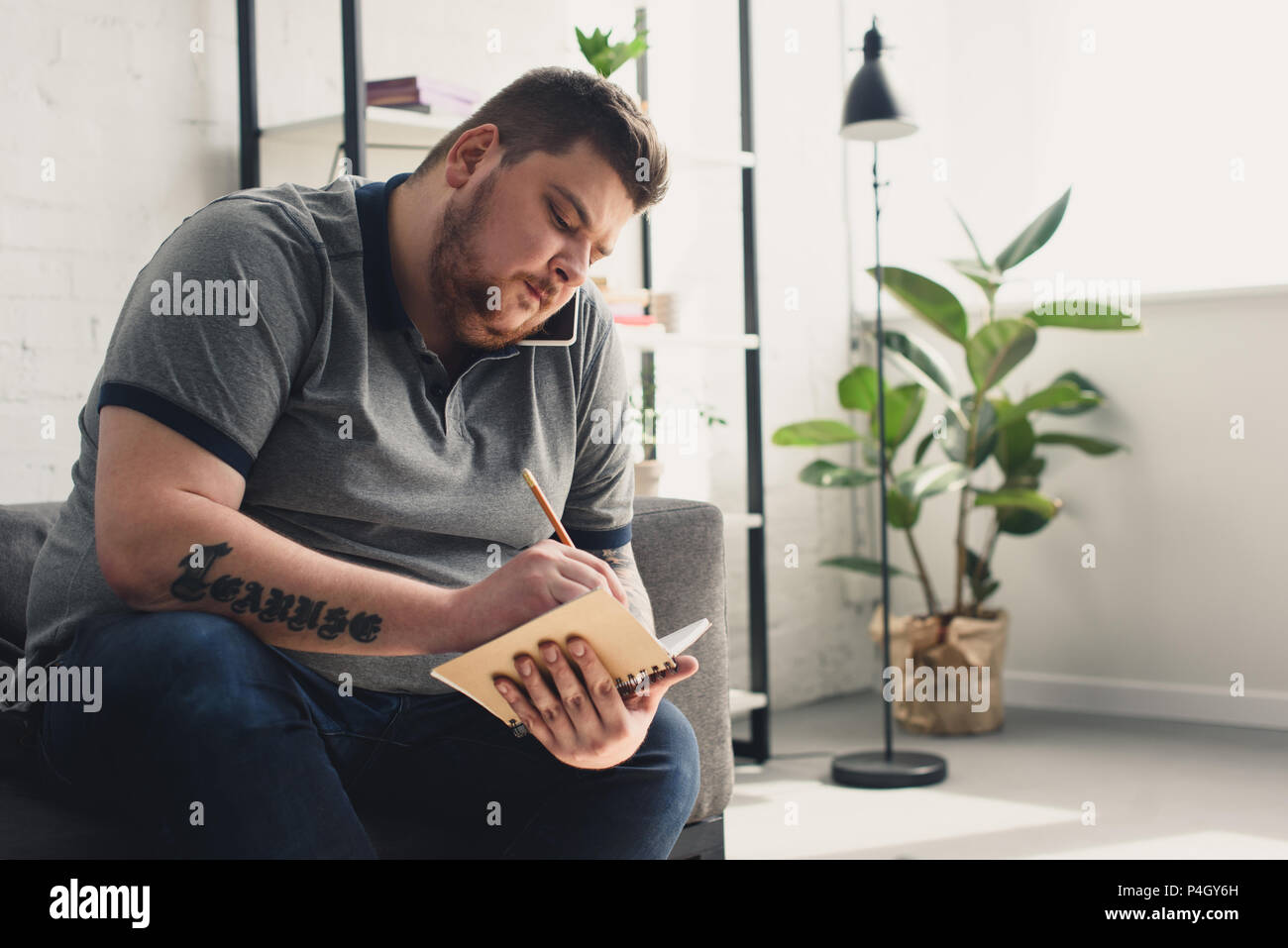 Handsome overweight man writing something to notebook on sofa at home ...