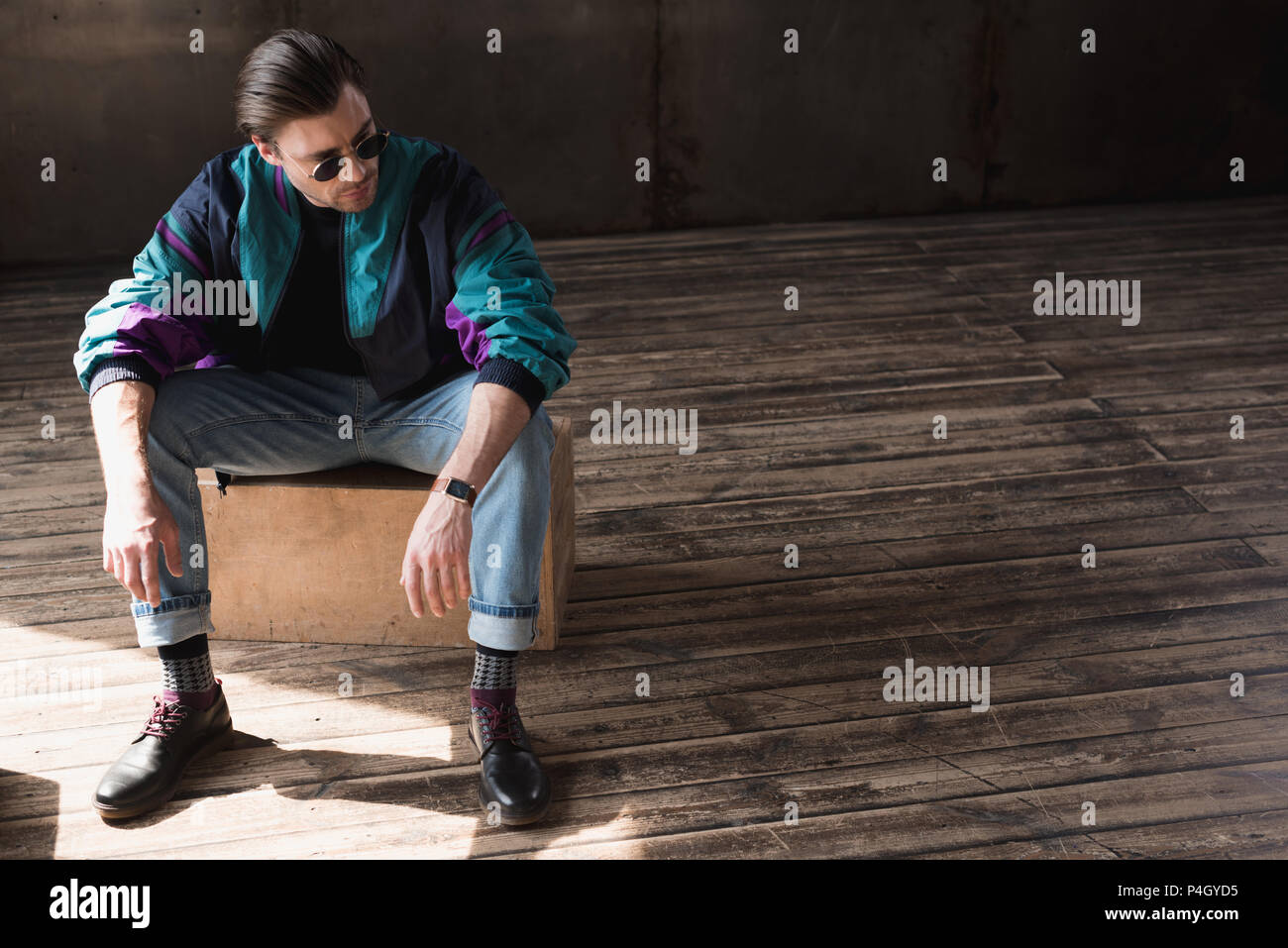 stylish young man in vintage windcheater sitting on wooden box in loft ...