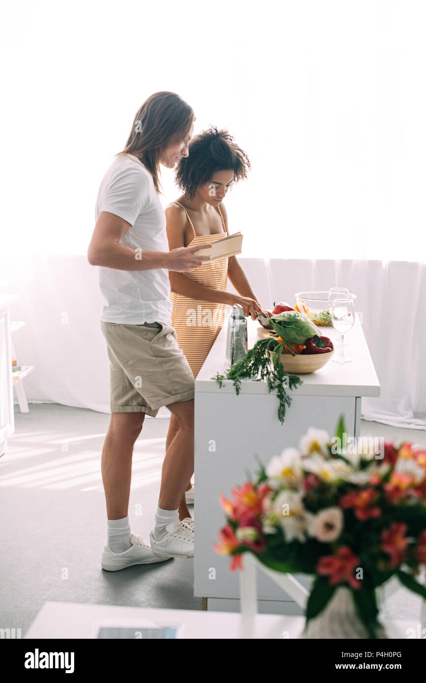 side view of multicultural couple with recipe in book cooking at ...