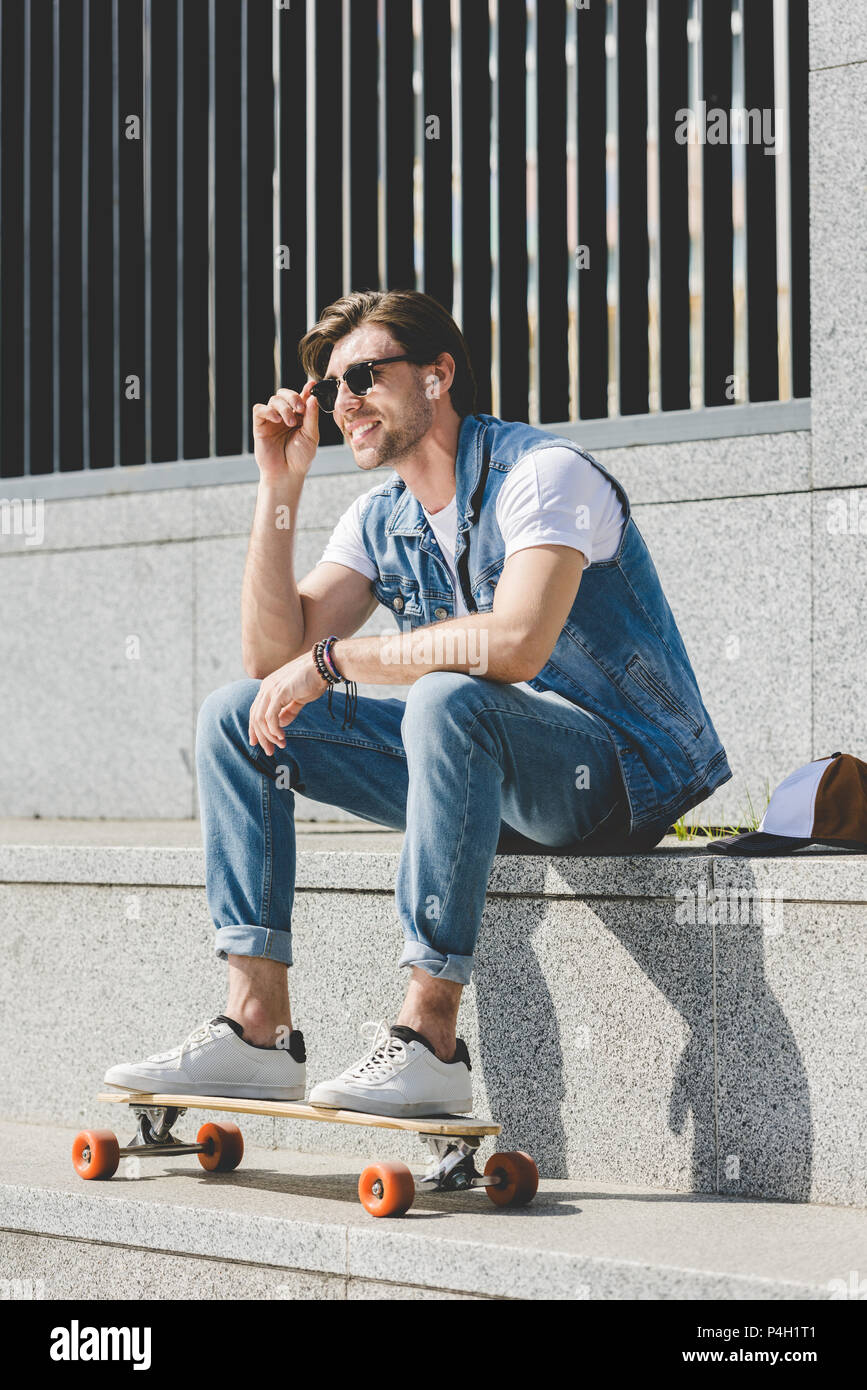 handsome young skater sitting on stairs with skateboard looking away ...