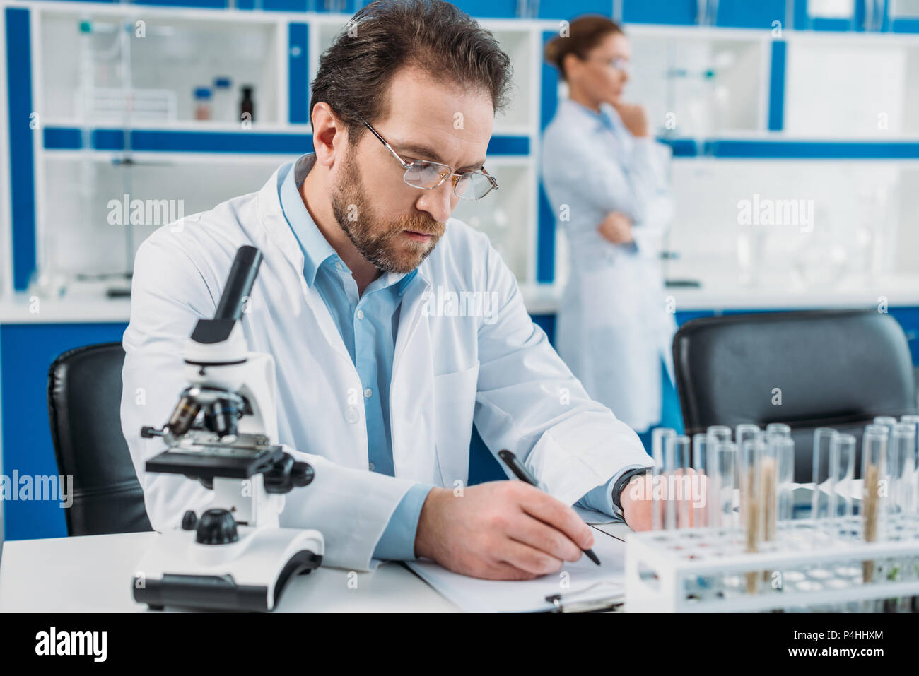 selective focus of scientist in white coat and eyeglasses making notes ...