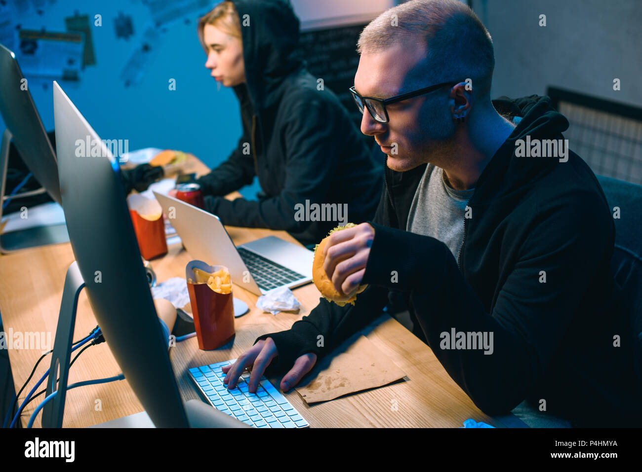 male hacker working on malware with accomplice and eating junk food ...