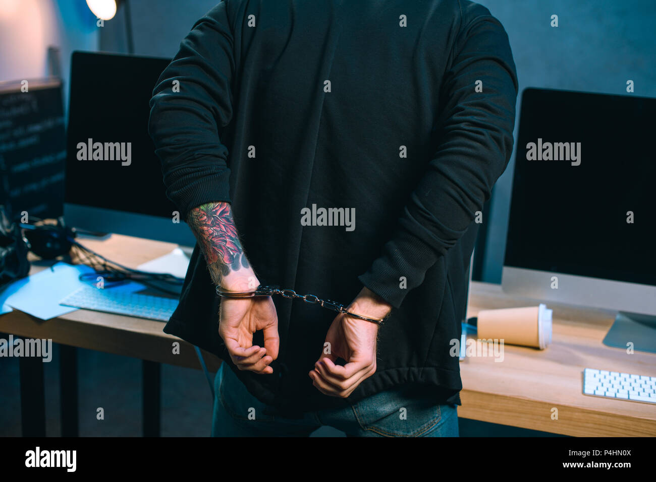 cropped shot of handcuffed hacker in hoodie in front of his work desk ...