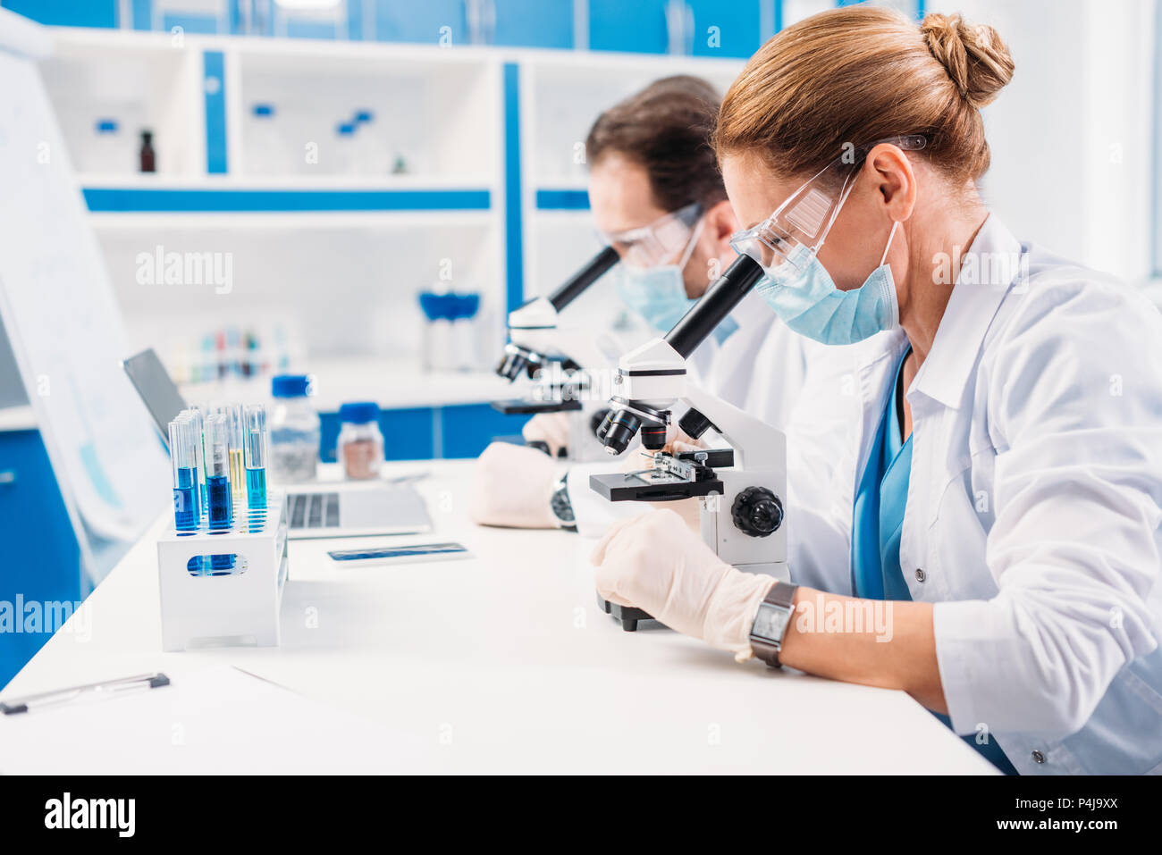 selective focus of scientists in medical masks and goggles looking through microscopes on ...