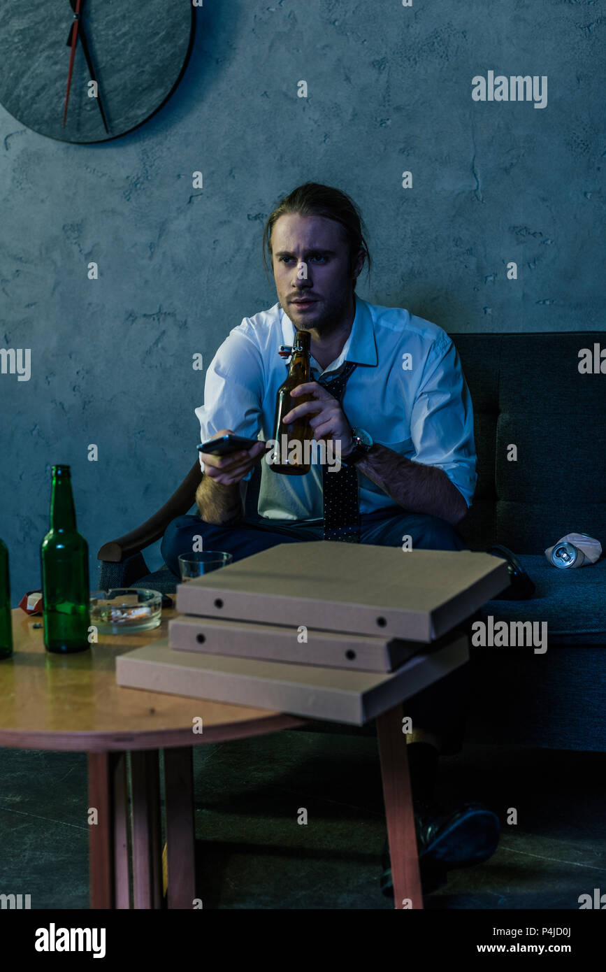 young alcohol addicted man in white shirt watching tv and drinking beer ...