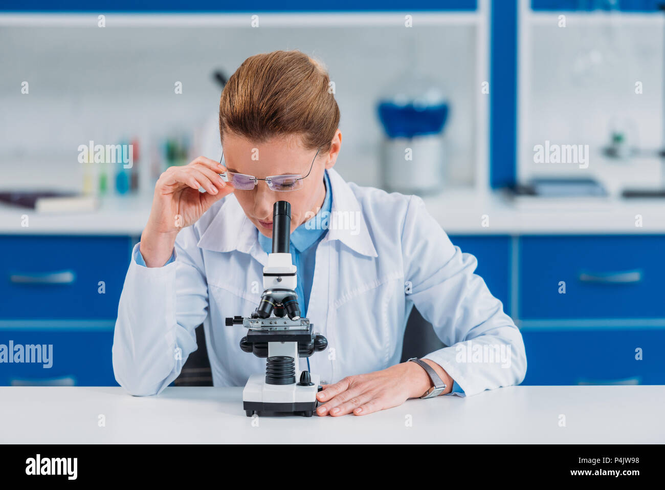 female scientist in eyeglasses looking on reagent through microscope in ...