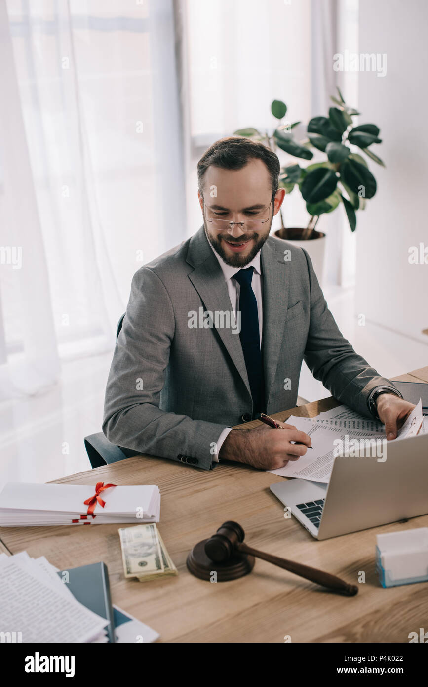 smiling lawyer in suit looking at bribe at workplace with laptop in ...