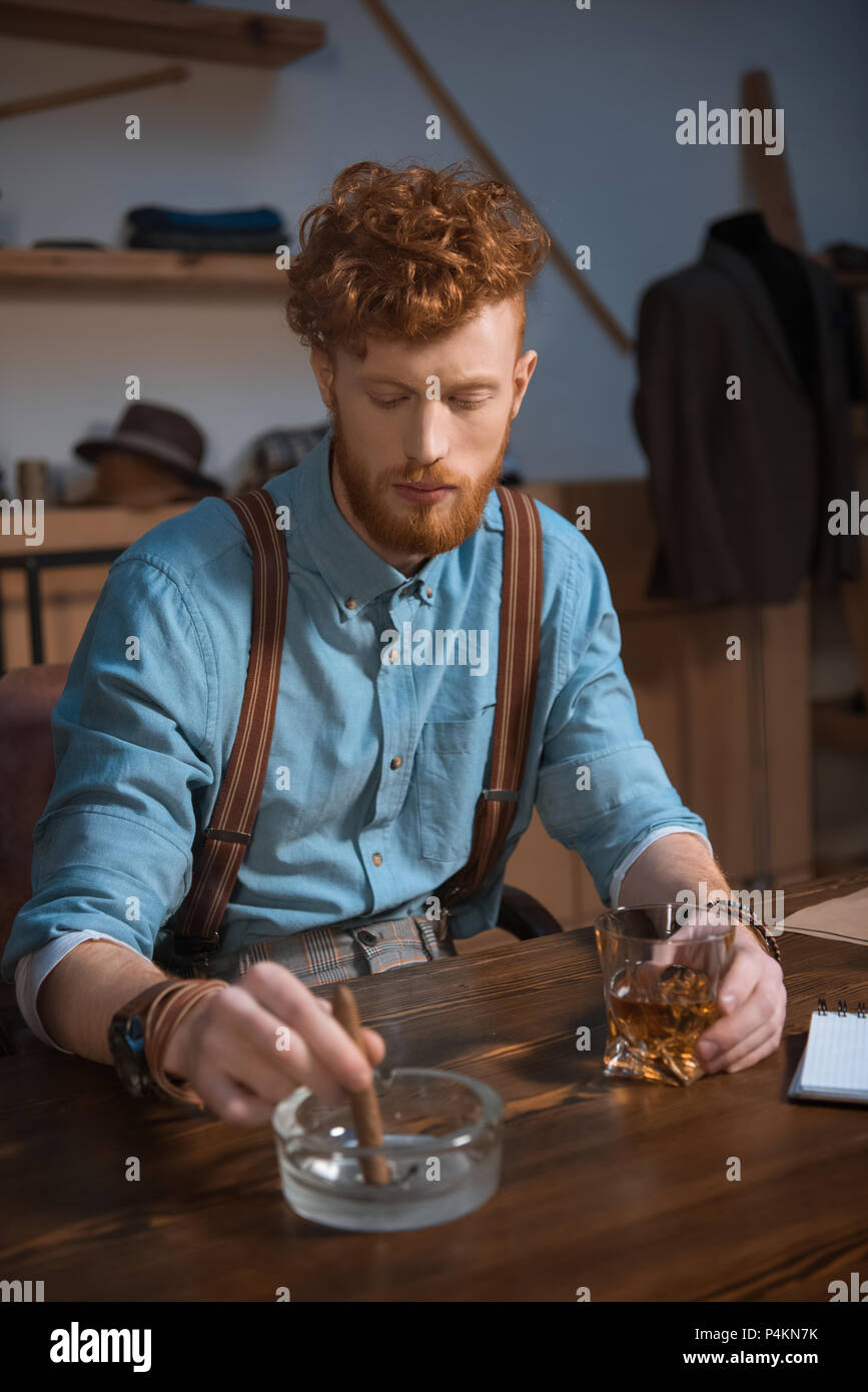 handsome young designer drinking whisky and smoking cigar at workplace ...