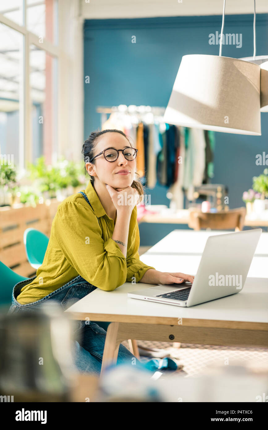 Portrait of fashion designer sitting at desk in her studio with laptop ...