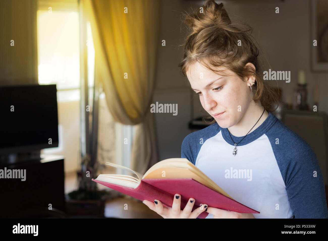 A teenager girl standing reading a book at home in the living room ...