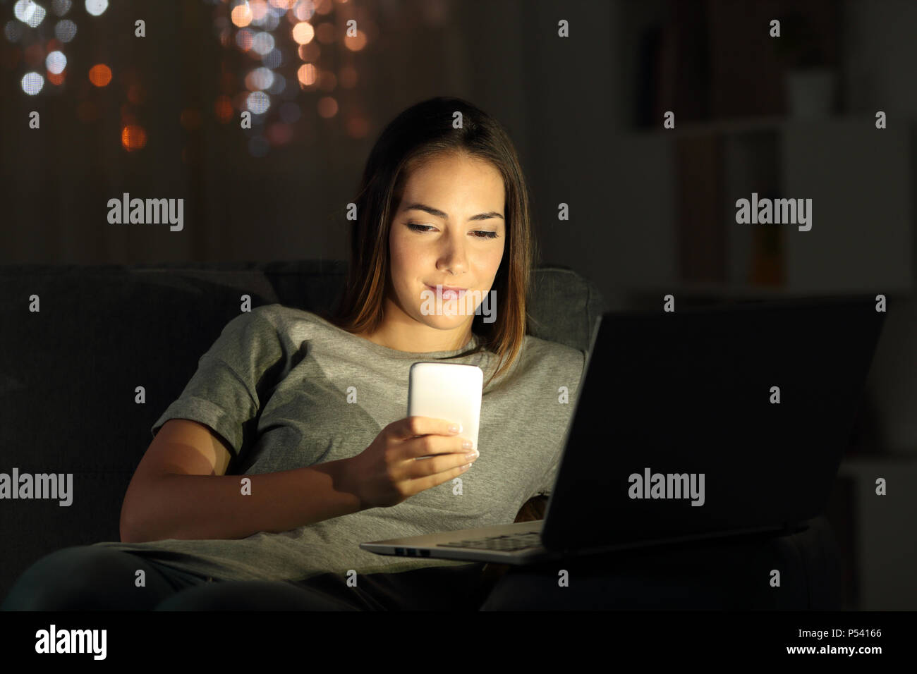Relaxed girl using a laptop and a phone in the night sitting on a couch ...