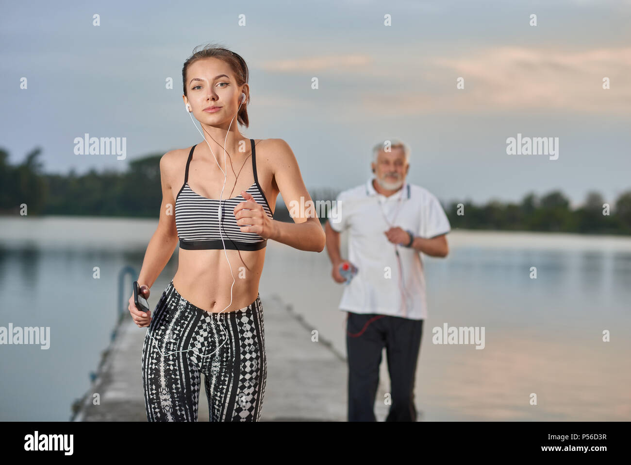 Sporty girl with fit figure running near lake in evening. Senior man ...